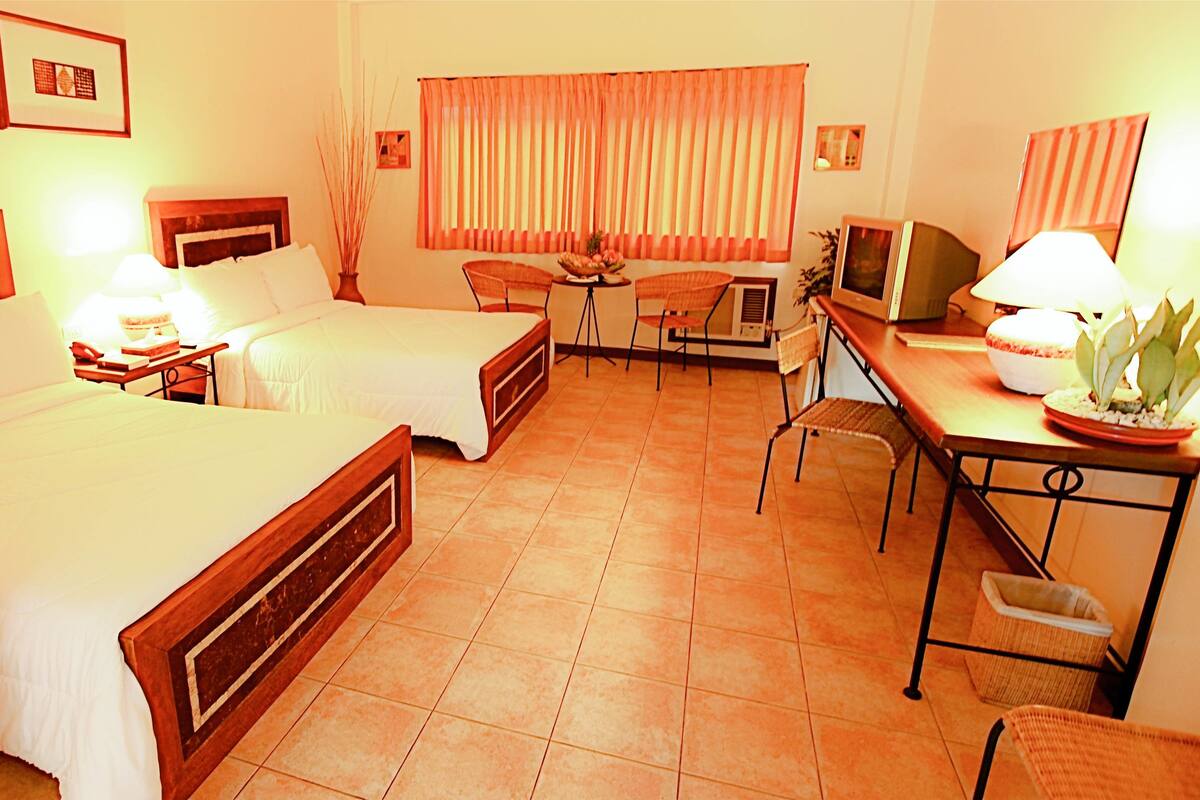 Quarto de hotel com duas camas, uma escrivaninha com computador, uma cadeira e uma televisão.