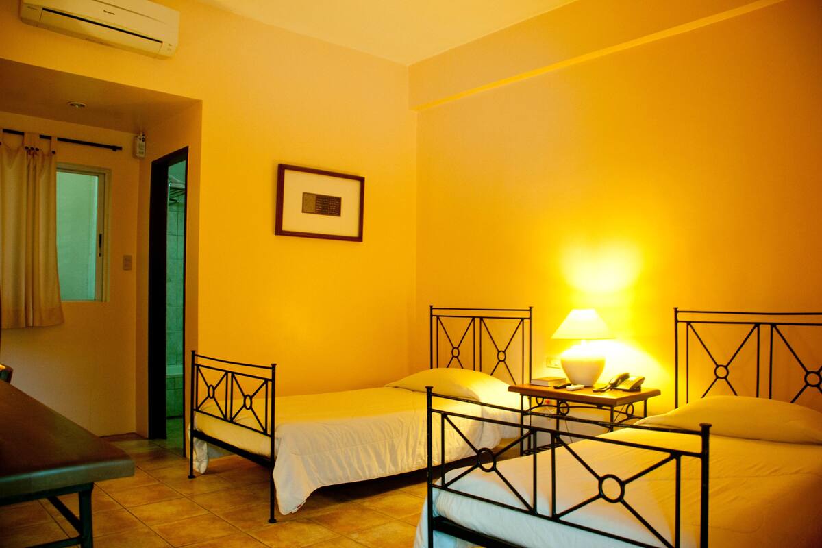 Quarto de hotel com duas camas de solteiro, uma mesa de cabeceira com abajur, um quadro na parede e um aparelho de ar condicionado.