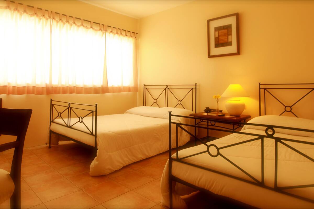 Quarto com duas camas de solteiro, uma escrivaninha, uma cadeira e um abajur.