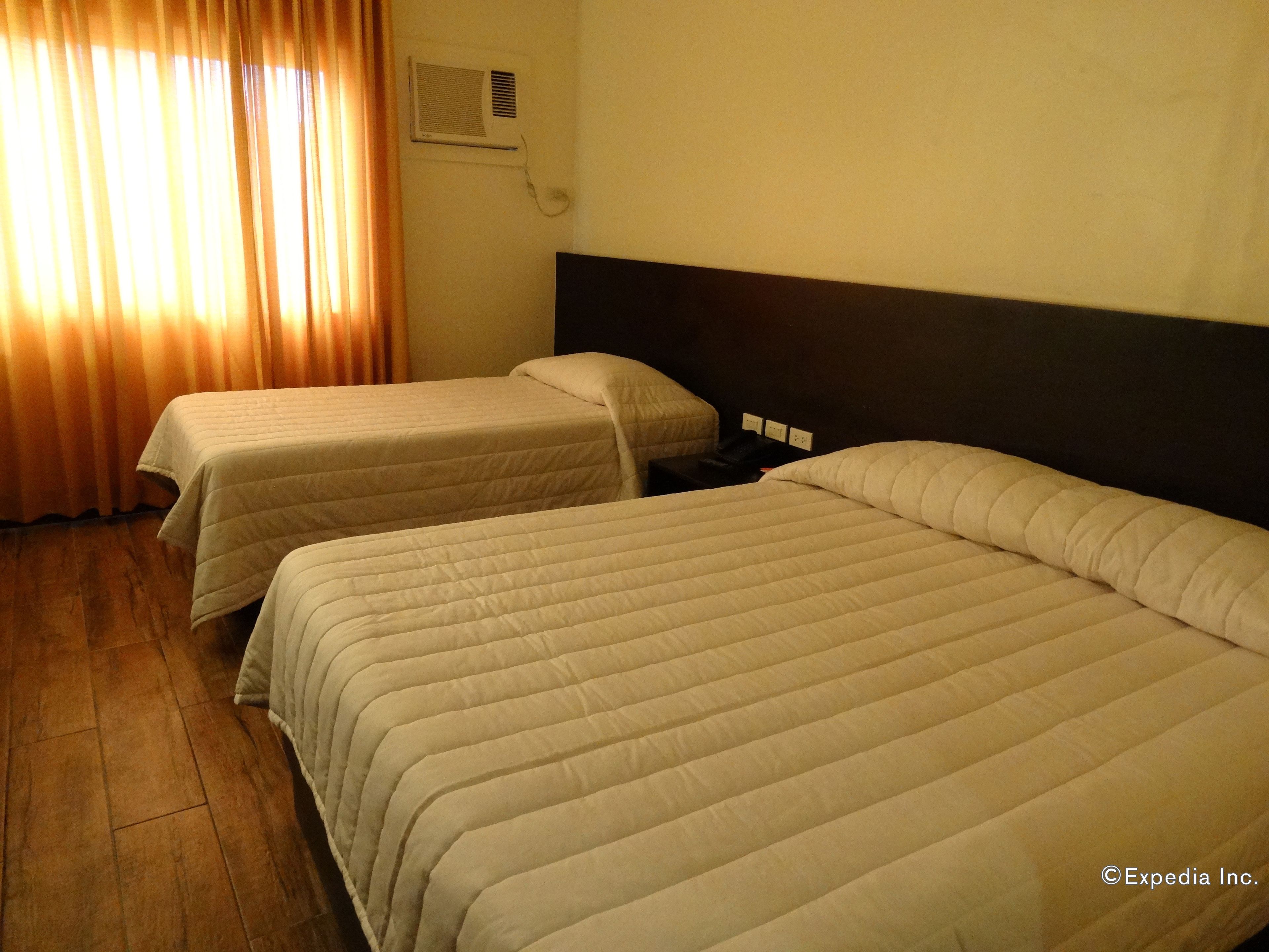 Duas camas de solteiro com cabeceiras, chão de madeira, janela com cortinas e ar condicionado fixado na parede.