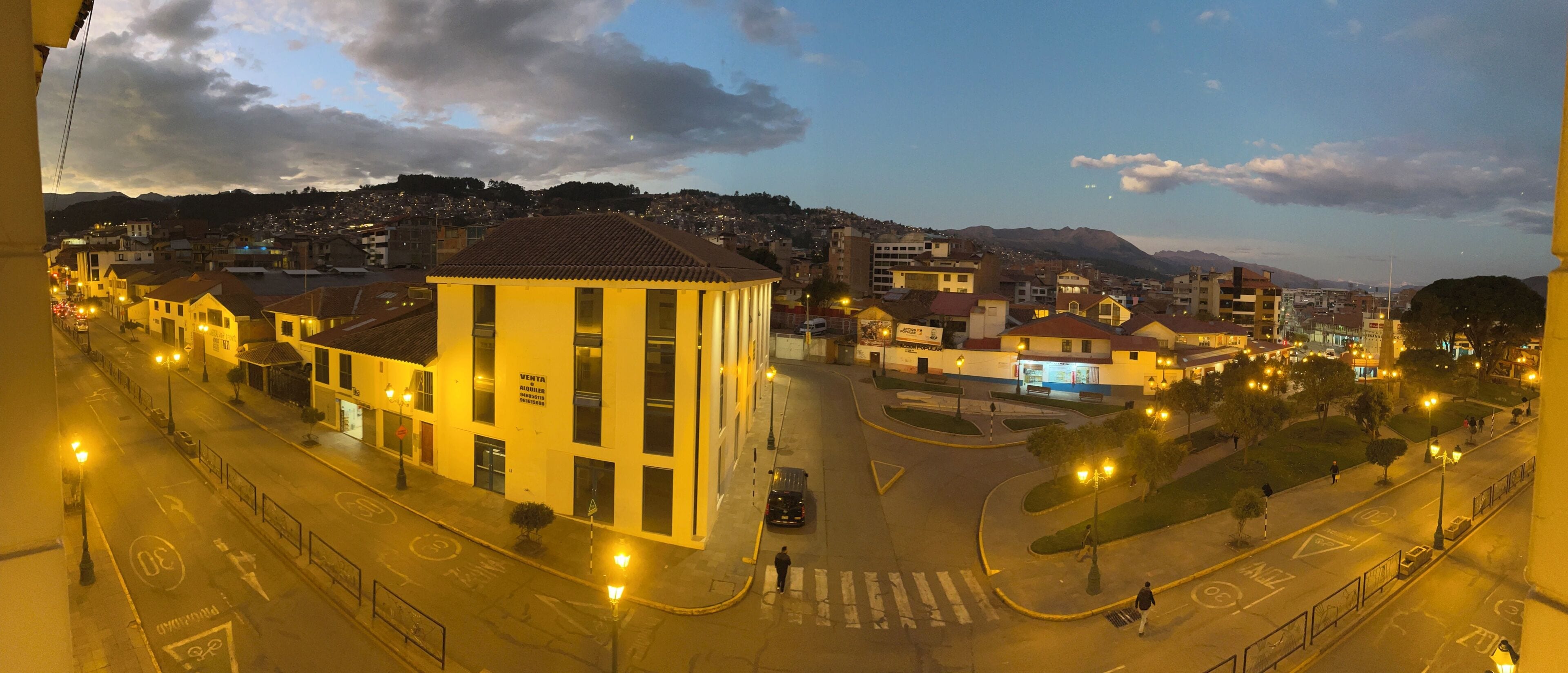 Vista urbana ao entardecer, com edifícios, ruas e uma faixa de pedestres.