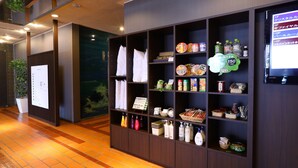 Uma área de recepção de hotel com um balcão de madeira, um ecrã digital a mostrar tarifas de quartos e estantes com produtos de higiene e snacks.