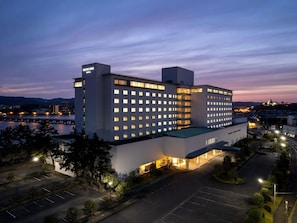 Um hotel de vários andares com design moderno, que apresenta um amplo estacionamento e vista para um rio e montanhas ao fundo.