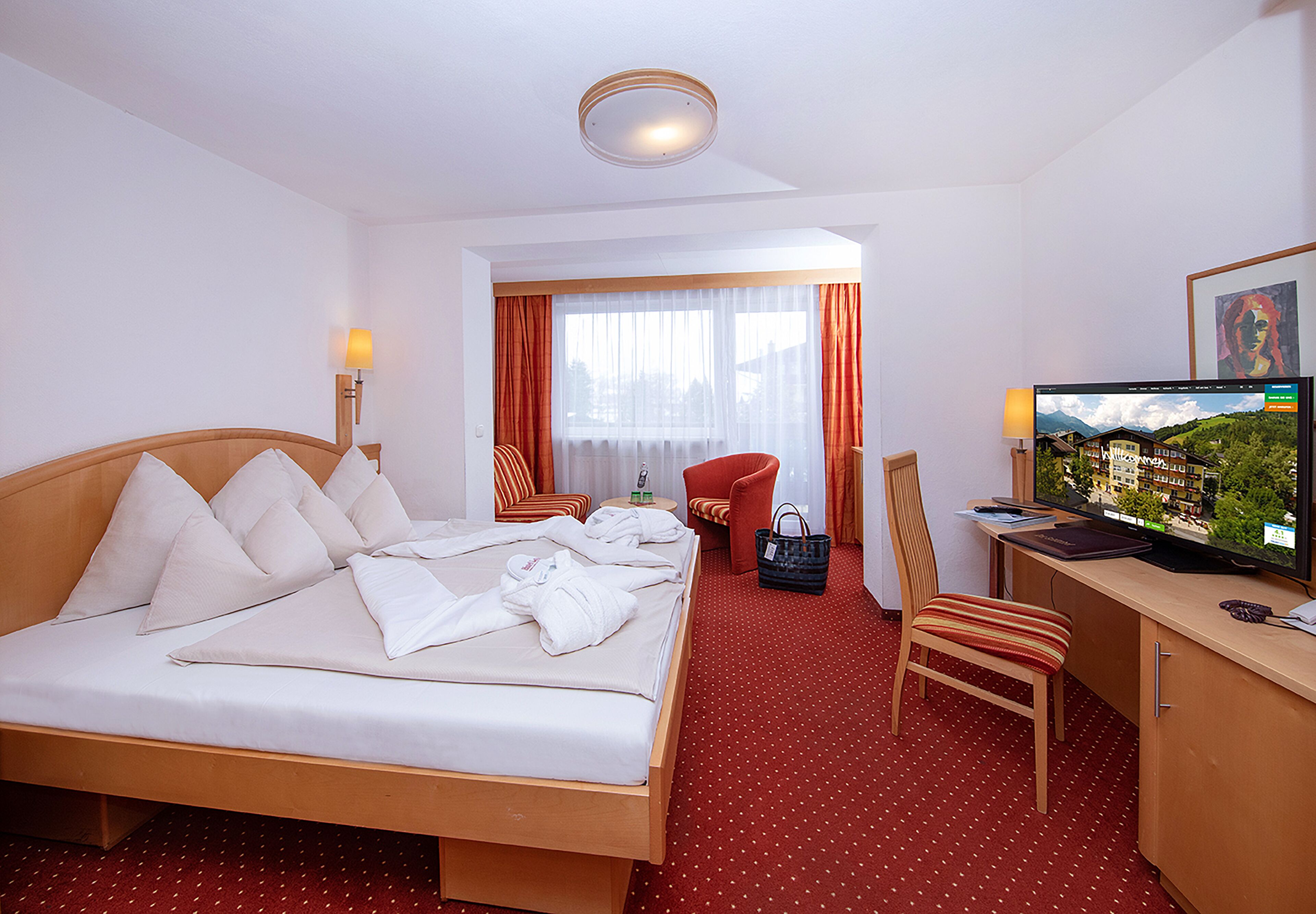 Quarto de hotel com uma cama, escrivaninha com computador, cadeira, televisão e janela com cortinas.