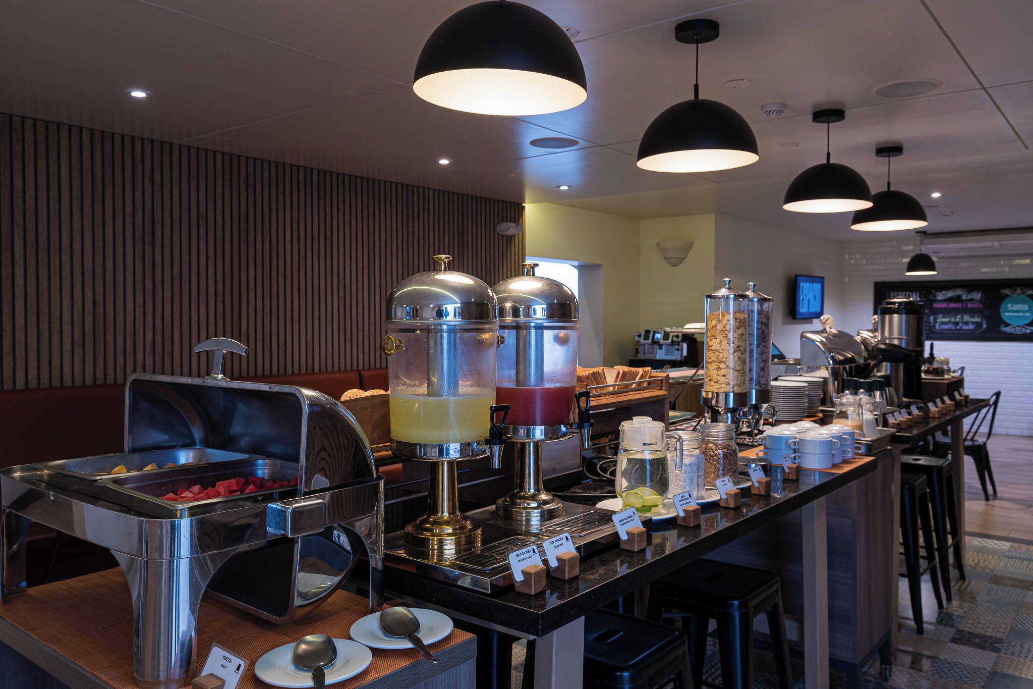 Disposição de buffet com diversos dispensadores de comida e bebida, uma máquina de café e uma área de estar com cadeiras.