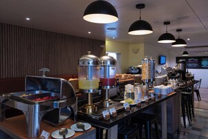 Disposição de buffet com diversos dispensadores de comida e bebida, uma máquina de café e uma área de estar com cadeiras.
