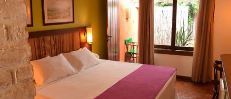 Quarto de hotel com cama, cabeceira de madeira, mesa de cabeceira com luminária, janela com cortinas e vista para o exterior.