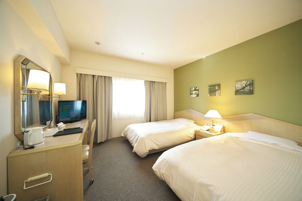 Quarto de hotel com duas camas, uma escrivaninha com computador, um espelho e uma janela com cortinas.