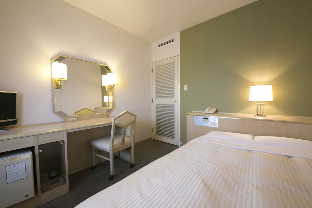 Quarto de hotel com cama, escrivaninha, cadeira e duas luminárias fixadas na parede.