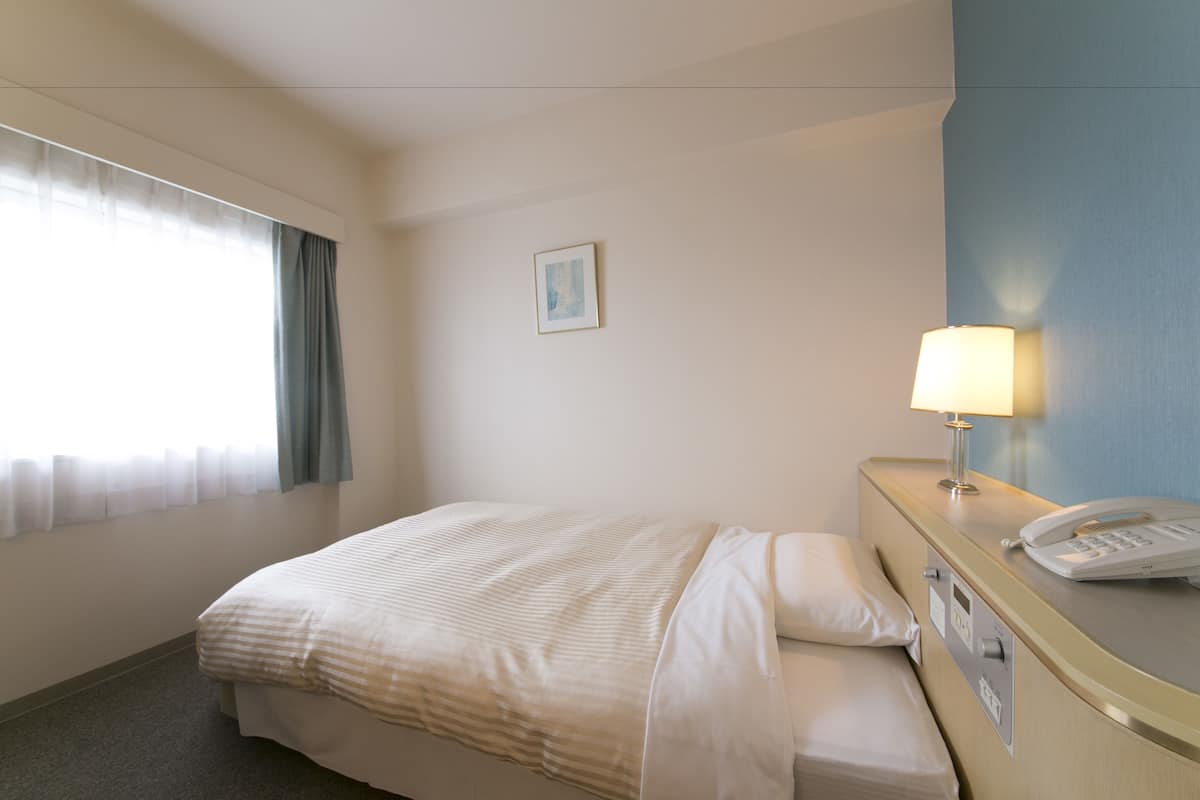 Quarto de hotel com cama, mesa de cabeceira com abajur, telefone e janela com cortinas.