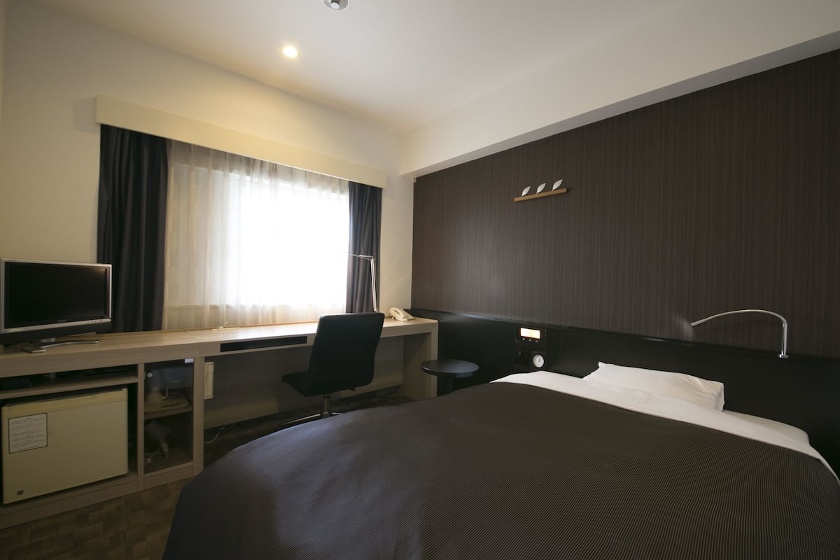 Quarto de hotel com cama, escrivaninha, cadeira, televisão e um pequeno armário.