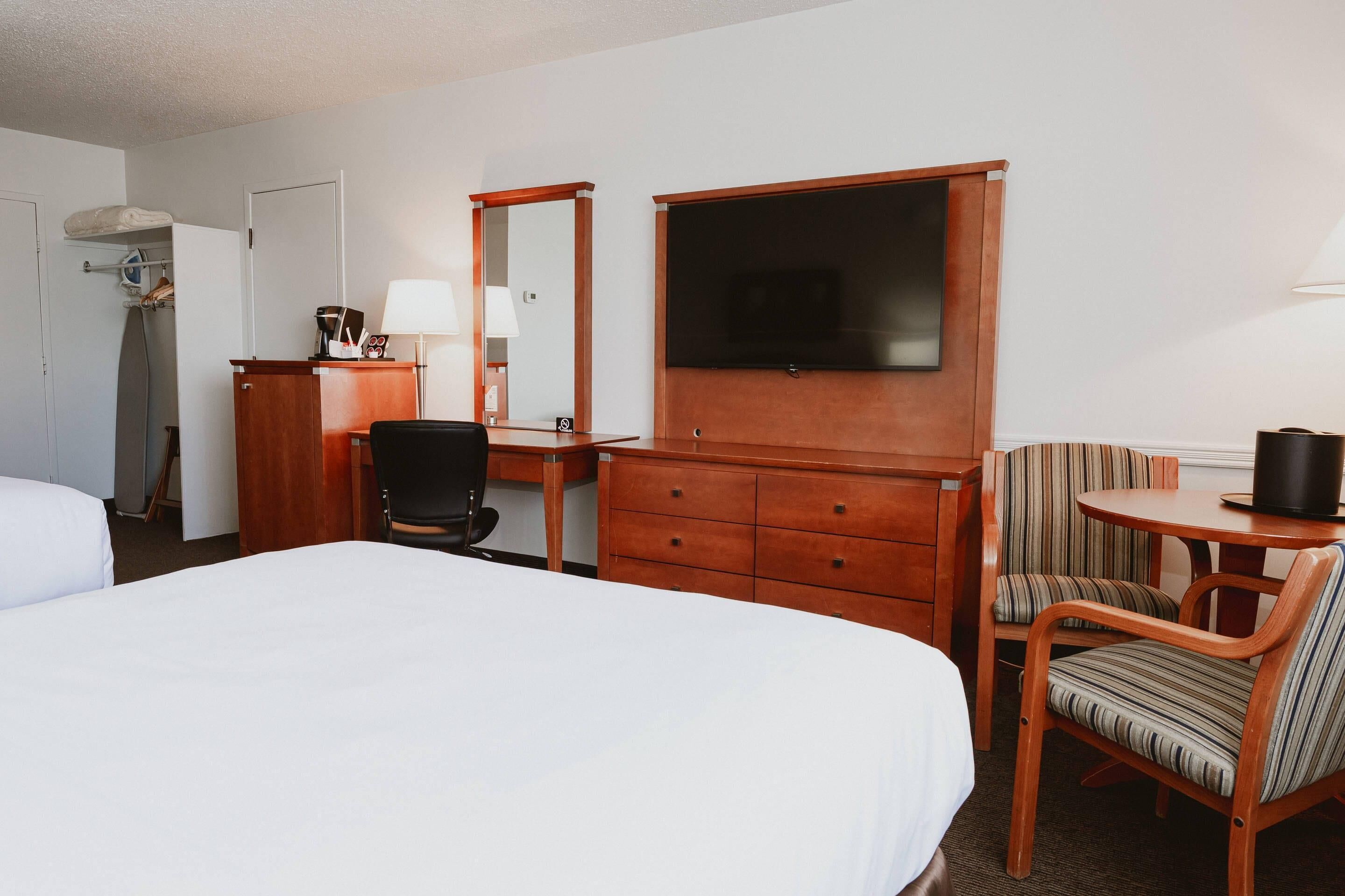 Quarto de hotel com uma cama grande, uma escrivaninha com cadeira, uma televisão, um espelho e uma pequena mesa com cadeira.