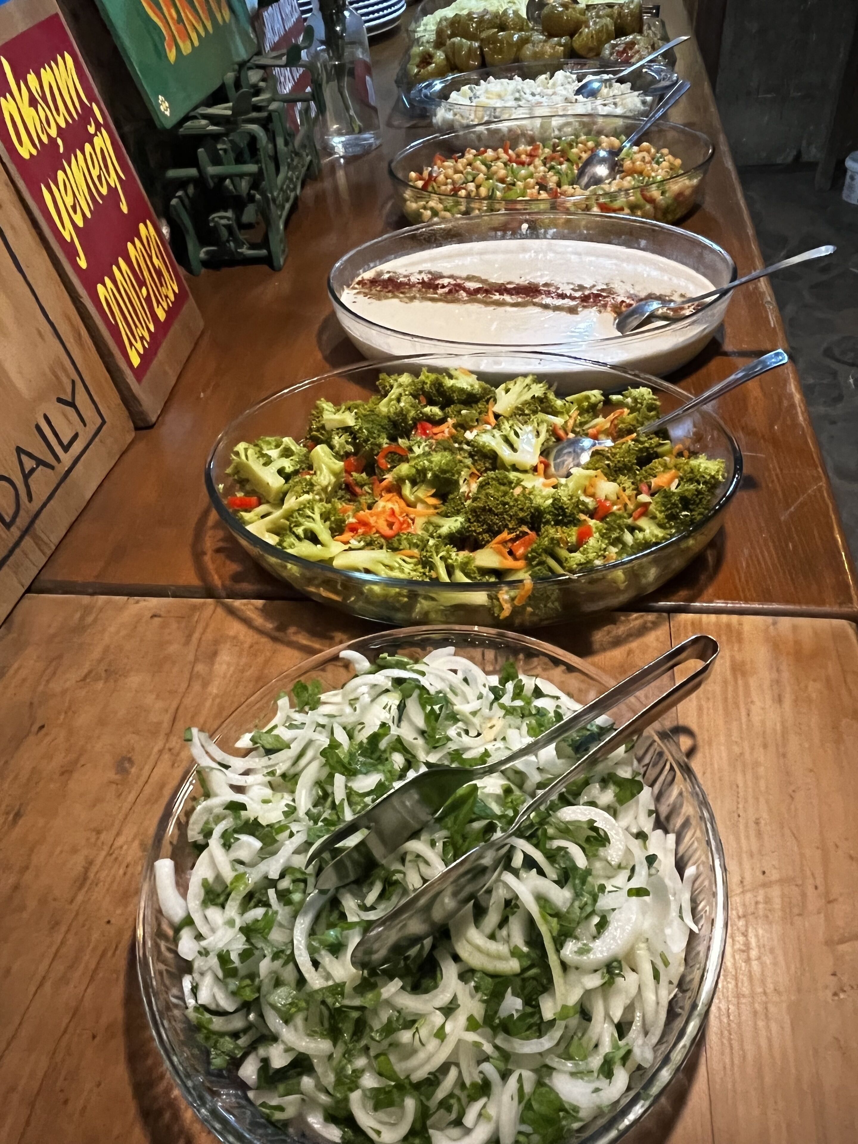 Mesa de buffet com diversos pratos, incluindo saladas, legumes e um letreiro com o menu diário.