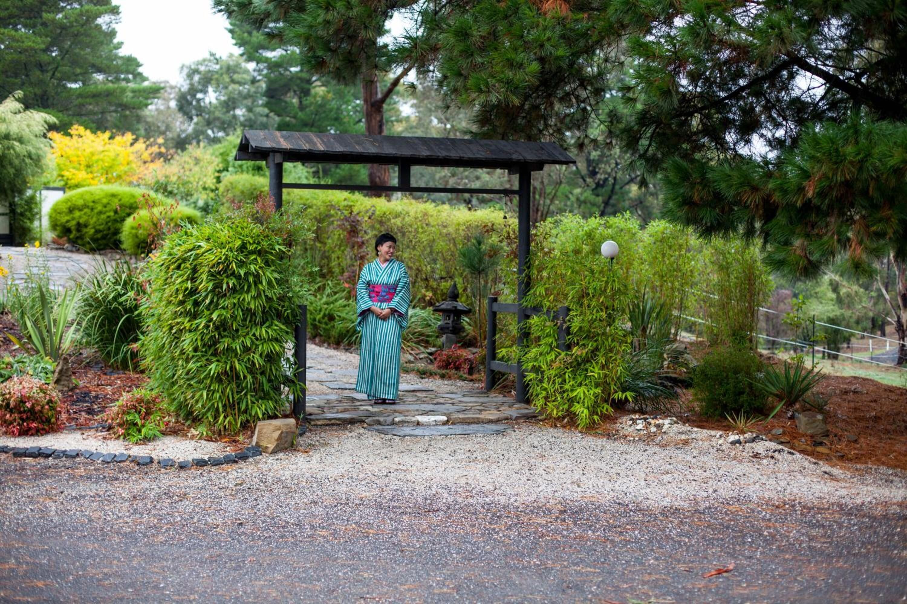 Uma pessoa em baixo de um gazebo de madeira num jardim com diversas plantas e árvores.