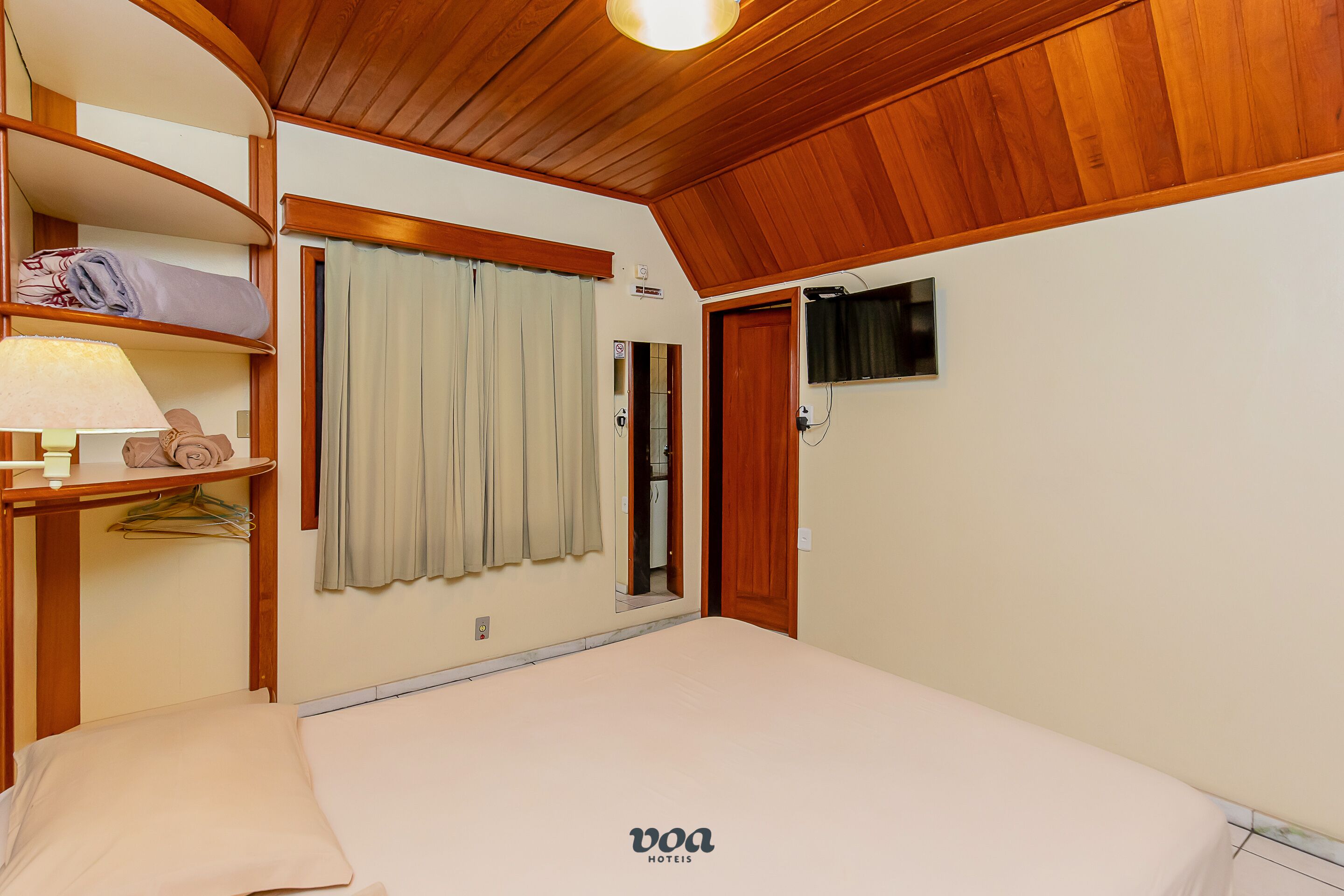 Quarto de hotel com cama, escrivaninha com luminária, guarda-roupa e janela com cortinas.