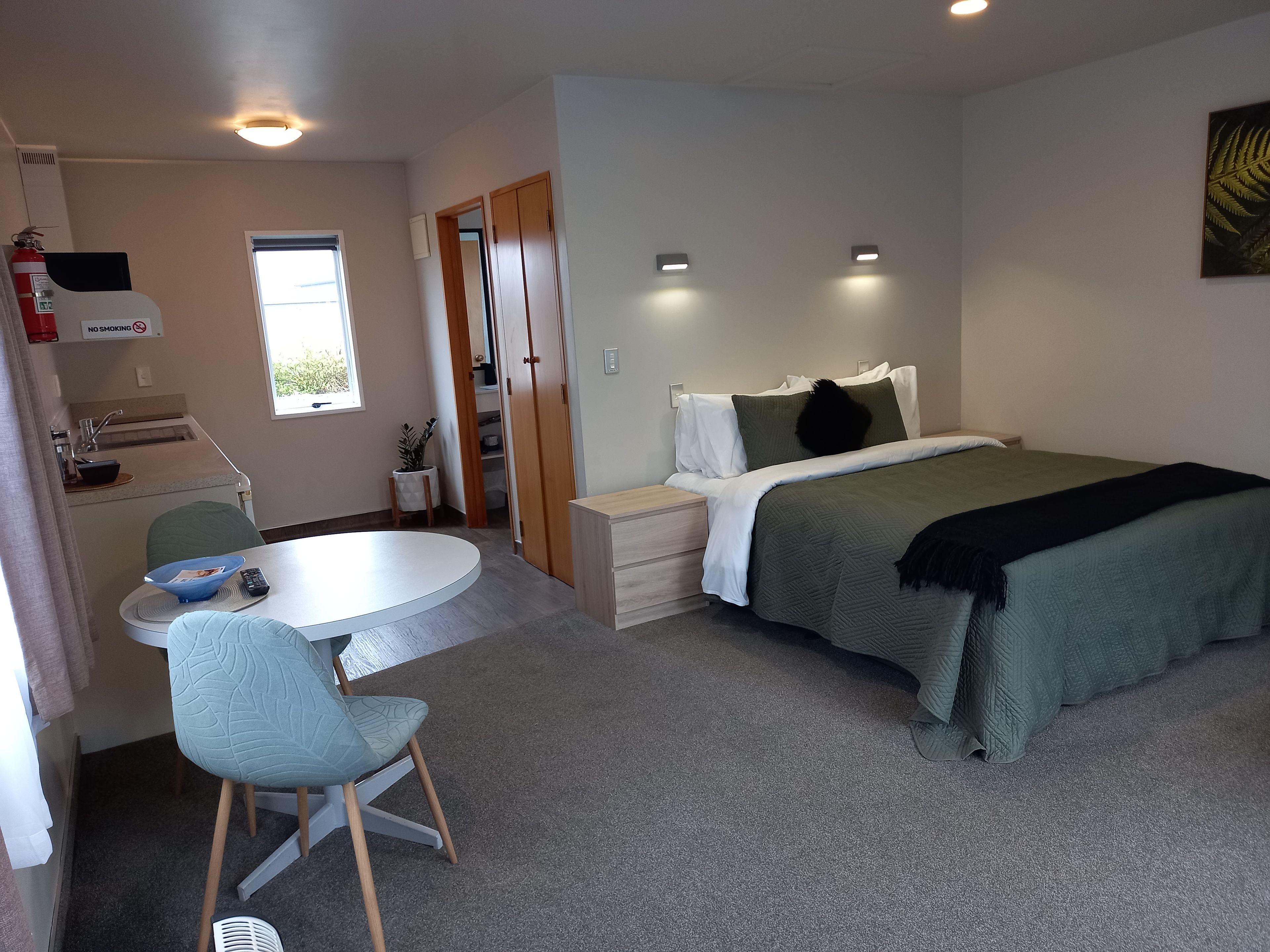 Quarto de hotel com cama, mesa de cabeceira, cadeira e uma pequena cozinha.