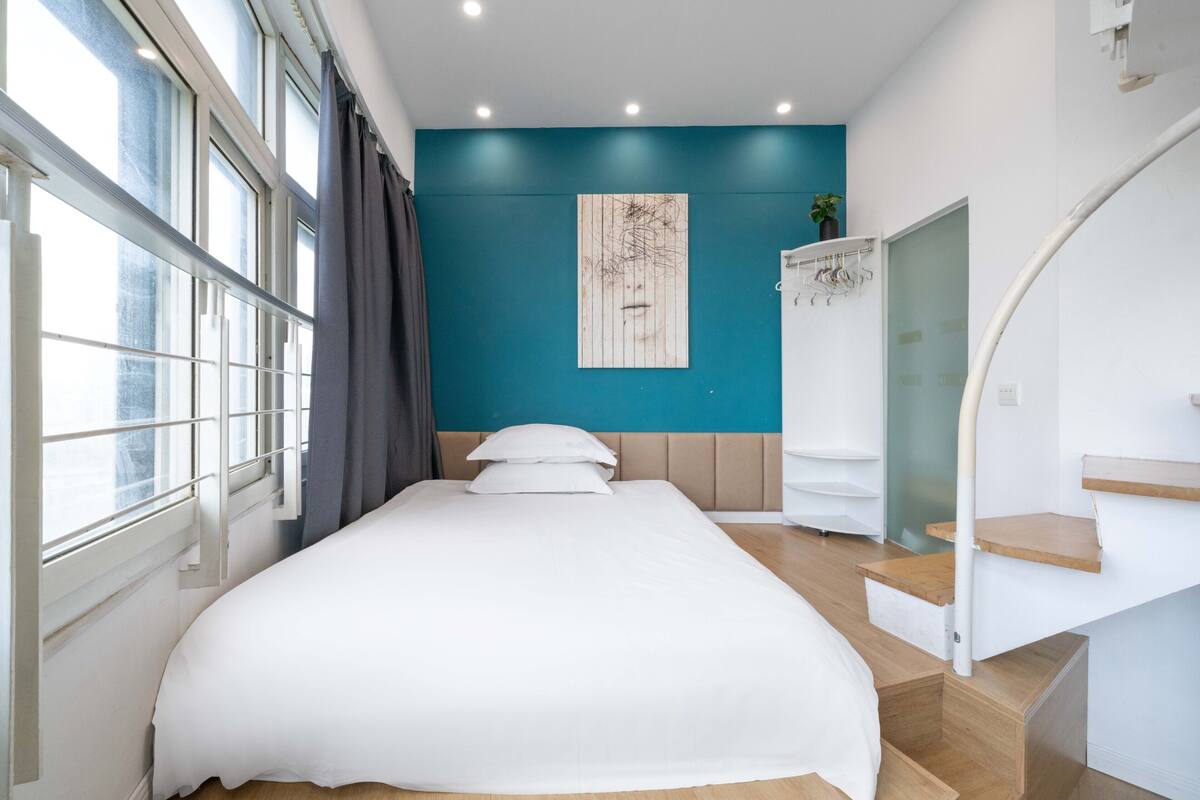 Quarto de hotel moderno com cama, janela com cortinas, parede em tom de verde-água, piso de madeira e escada.