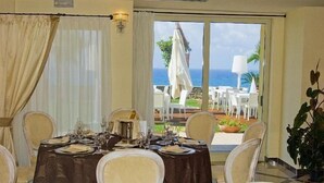 Uma sala de jantar com mesa posta para seis pessoas, com vista para uma área com piscina, guarda-sóis e cadeiras.