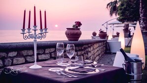 Mesa posta para refeição, com velas, copos de vinho e uma garrafa de champanhe, com vista para um pôr do sol sobre a água.