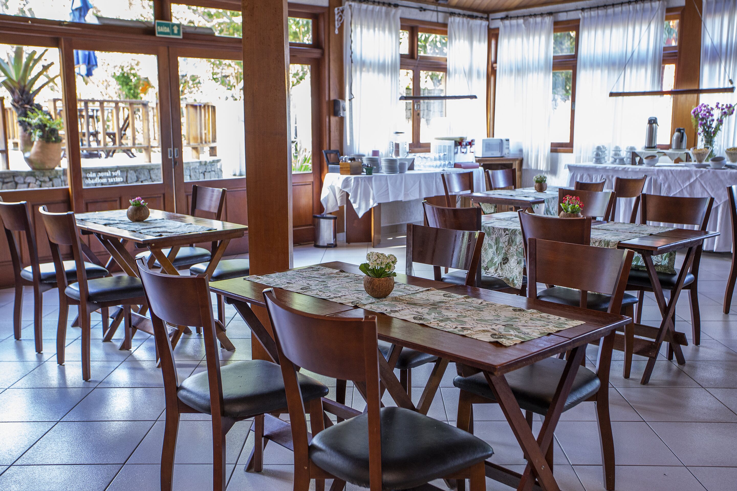 Restaurante com várias mesas e cadeiras, janelas amplas com cortinas e vista para o exterior.