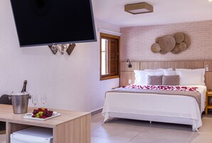 Quarto de hotel com uma cama, televisão fixada na parede, mesa de cabeceira com uma garrafa e copos, e parede de tijolos com pratos decorativos.