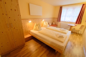 Quarto de hotel com cama de madeira, uma mesa pequena, uma cadeira e janela com cortinas.