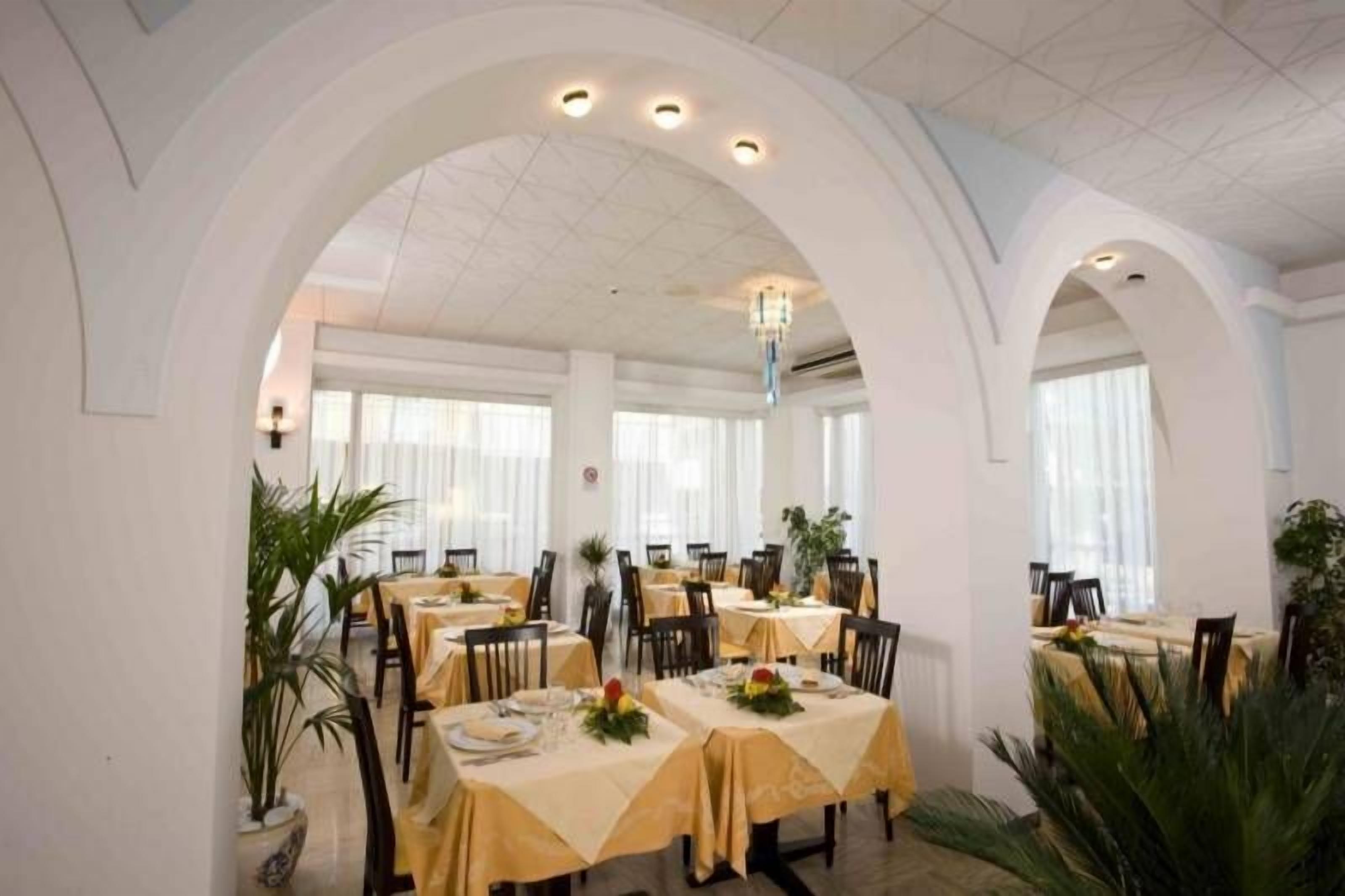 Uma sala de jantar com mesas preparadas para o serviço, paredes brancas e um lustre.
