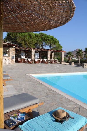 Área da piscina com espreguiçadeiras, uma mesa com chapéu e óculos de sol, e uma pérgula com assentos externos.