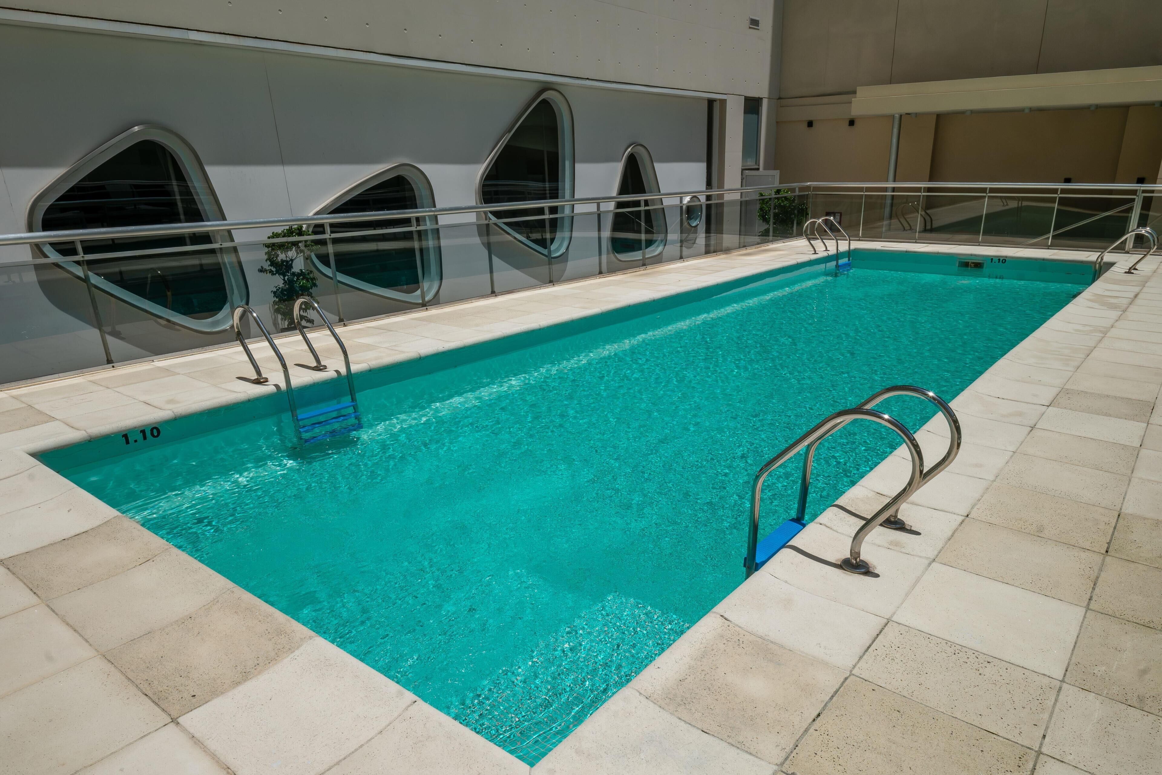Piscina retangular coberta com água azul cristalina, grades de metal e chão em mosaico.