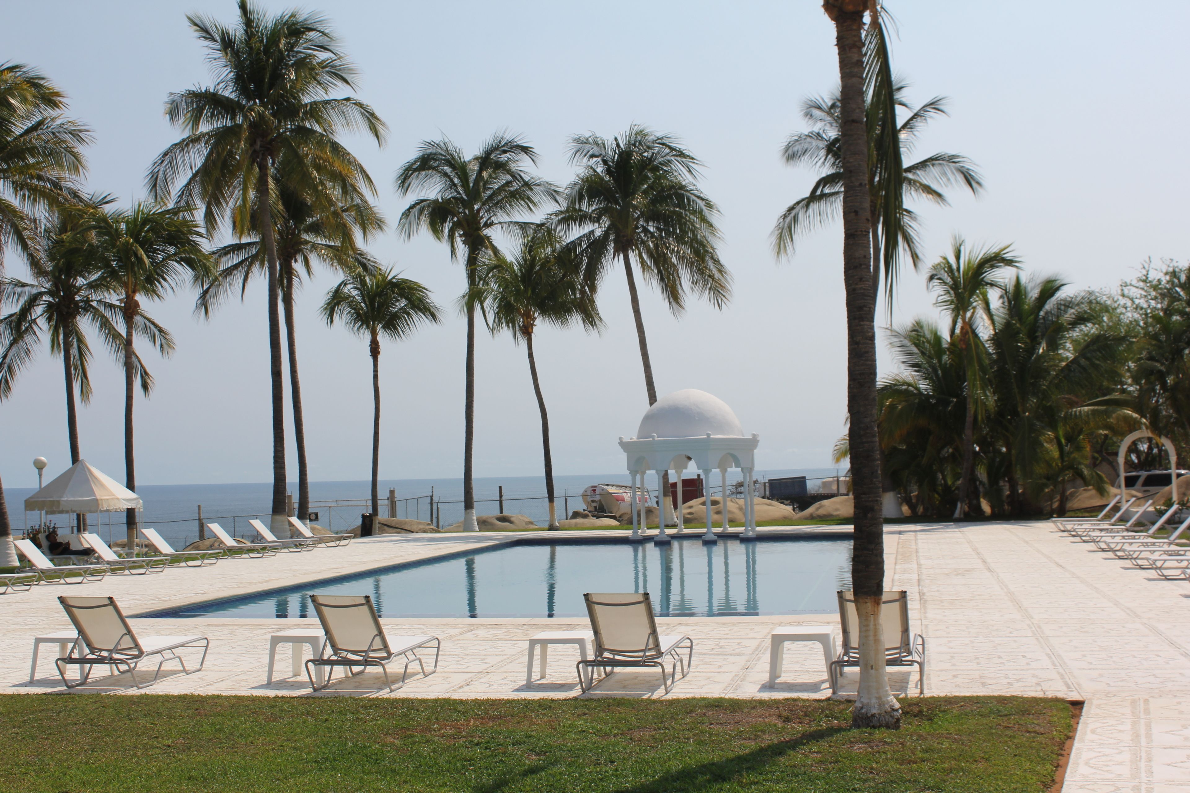 Área de piscina com espreguiçadeiras, palmeiras e um gazebo.