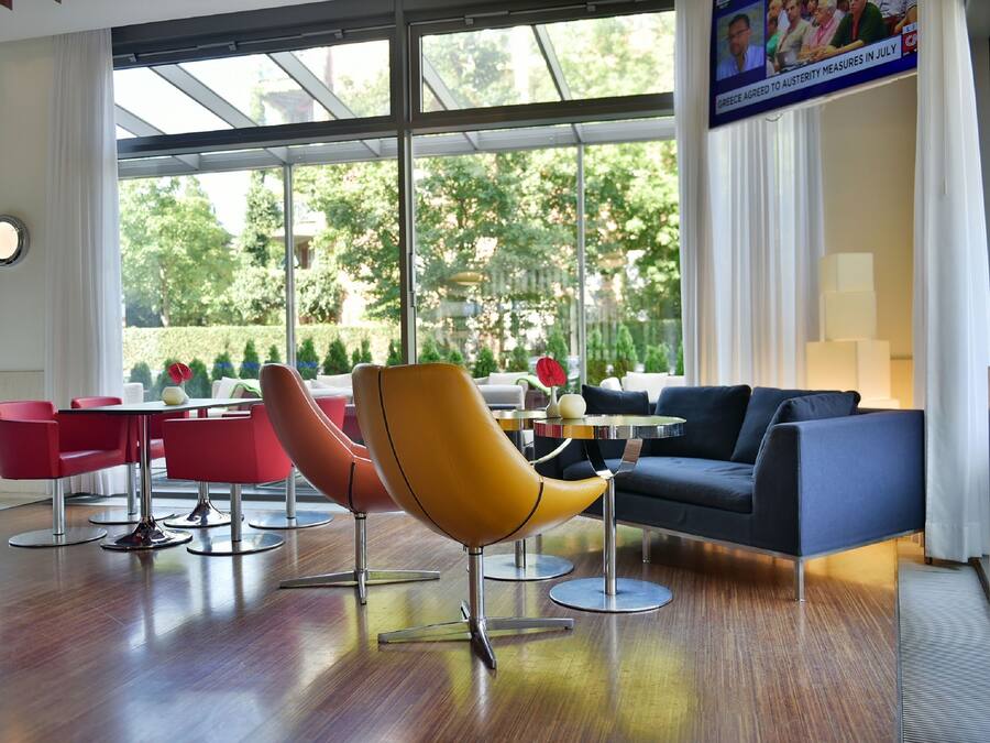 Um lobby moderno com piso de madeira, cadeiras vermelhas e amarelas, um sofá azul e janelas amplas com vista para o verde.
