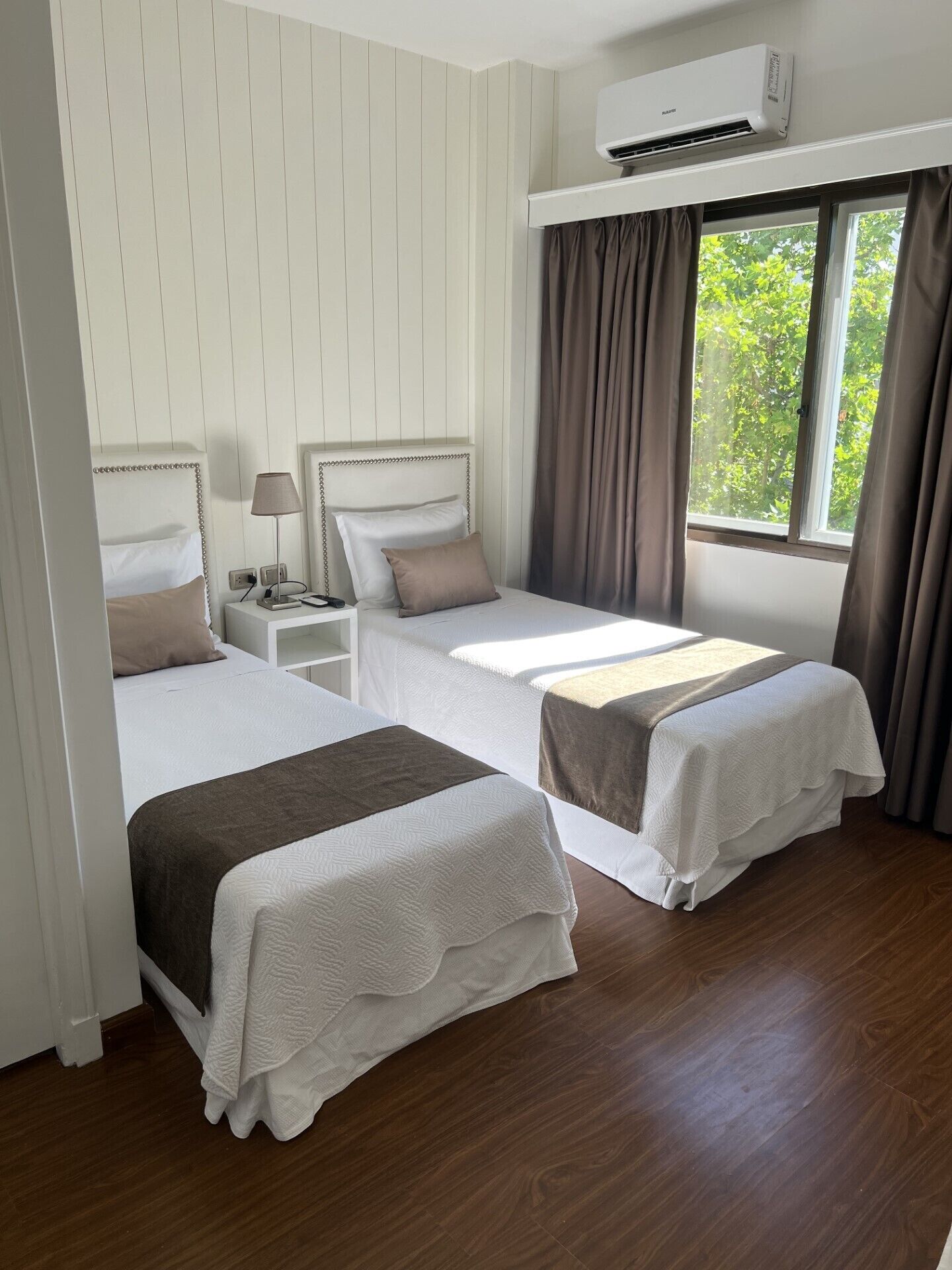 Quarto de hotel com duas camas, uma mesa de cabeceira, luminária, ar condicionado e janela com cortinas.