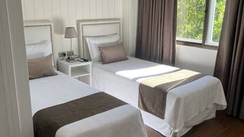 Quarto de hotel com duas camas, uma mesa de cabeceira, luminária, ar condicionado e janela com cortinas.
