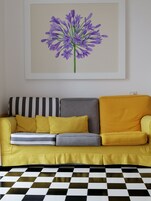 Um sofá amarelo com almofadas listradas e de cores sólidas, piso xadrez e uma obra de arte floral emoldurada acima.