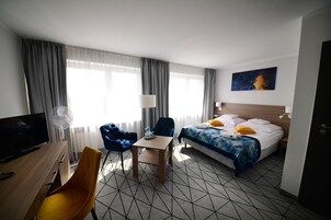 Quarto de hotel com cama, escrivaninha com televisão, cadeira, mesa e janela com cortinas.
