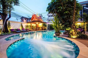 Piscina com espreguiçadeiras, edifício tradicional com telhado vermelho e vegetação ao redor.