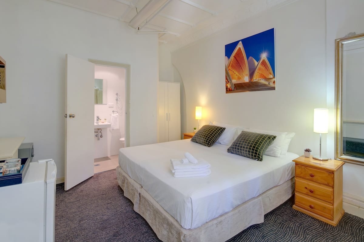 Quarto de hotel com cama, mesa de cabeceira, abajur, pintura de um edifício e banheiro visível por uma porta aberta.