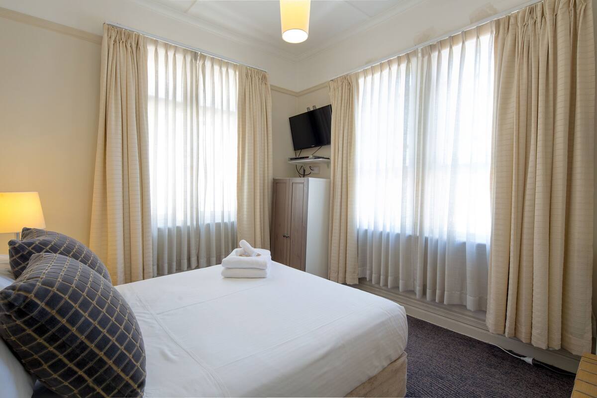 Quarto de hotel com cama, televisão, escrivaninha e radiador.