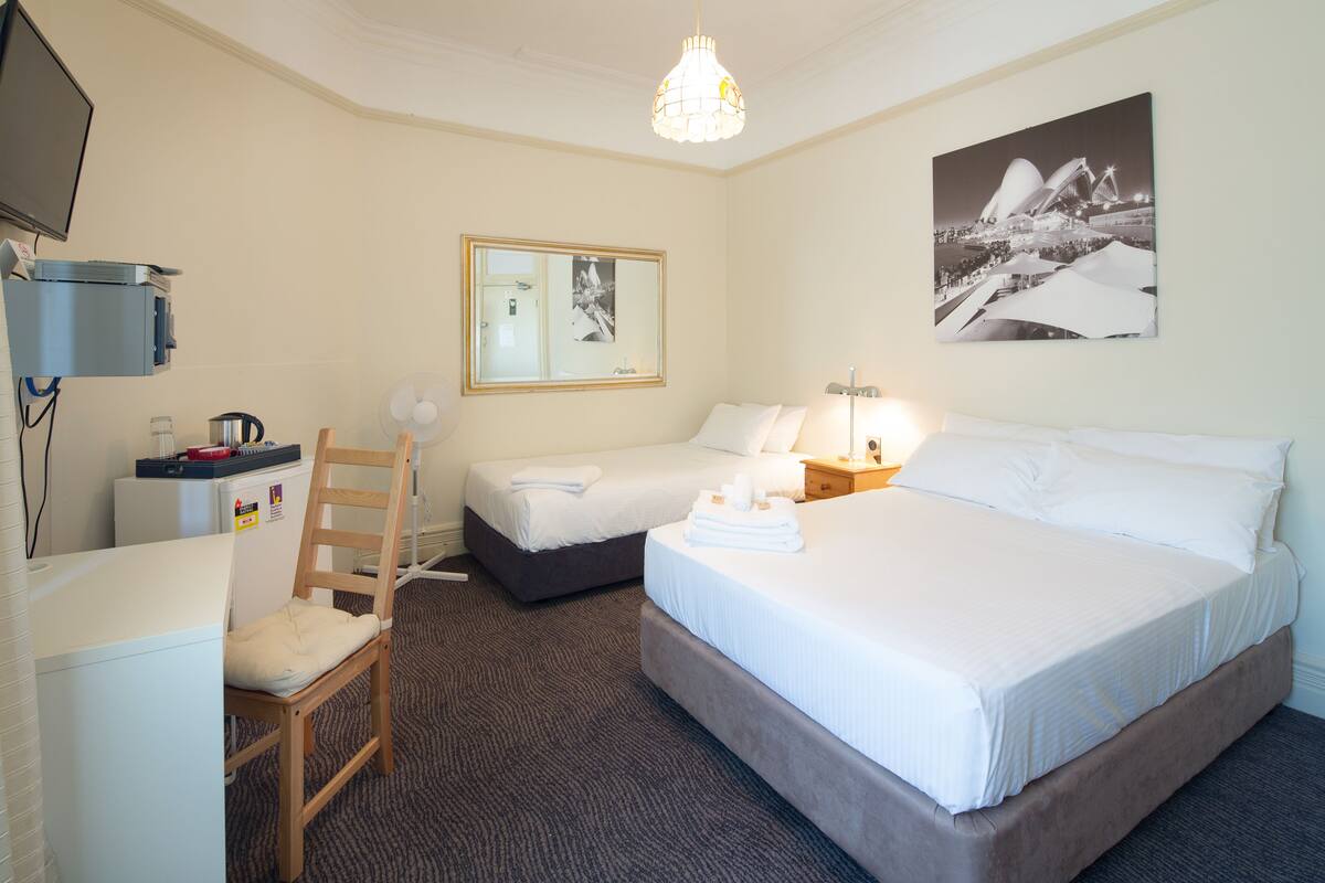 Quarto de hotel com uma cama grande, duas camas menores, uma cadeira, uma escrivaninha com um computador, um espelho e uma imagem da Sydney Opera House.