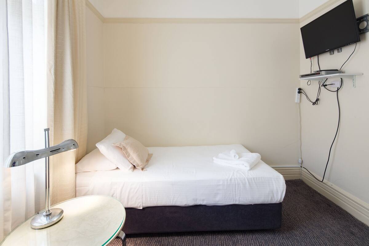 Quarto de hotel com uma cama de solteiro, mesa de cabeceira, televisão de ecrã plano na parede e tomada elétrica.