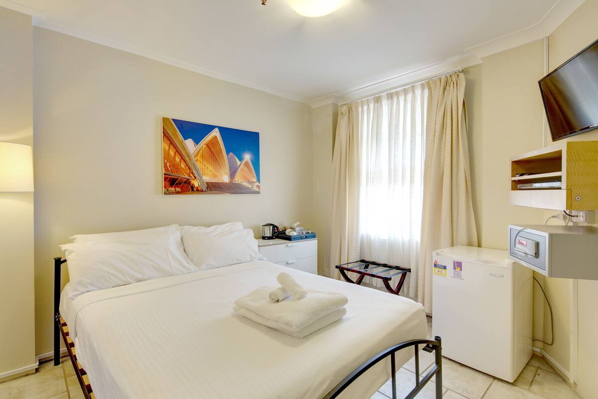 Quarto de hotel com cama, uma pequena mesa, geladeira, televisão e um quadro na parede.