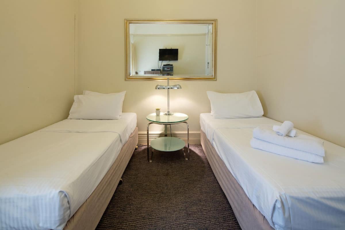 Quarto de hotel com duas camas de solteiro, uma mesinha redonda pequena, um abajur e um espelho na parede.