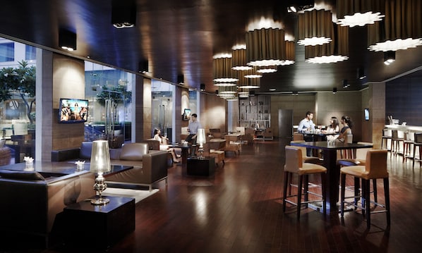 Um hall de hotel moderno com área de bar, assentos e uma tela de televisão.