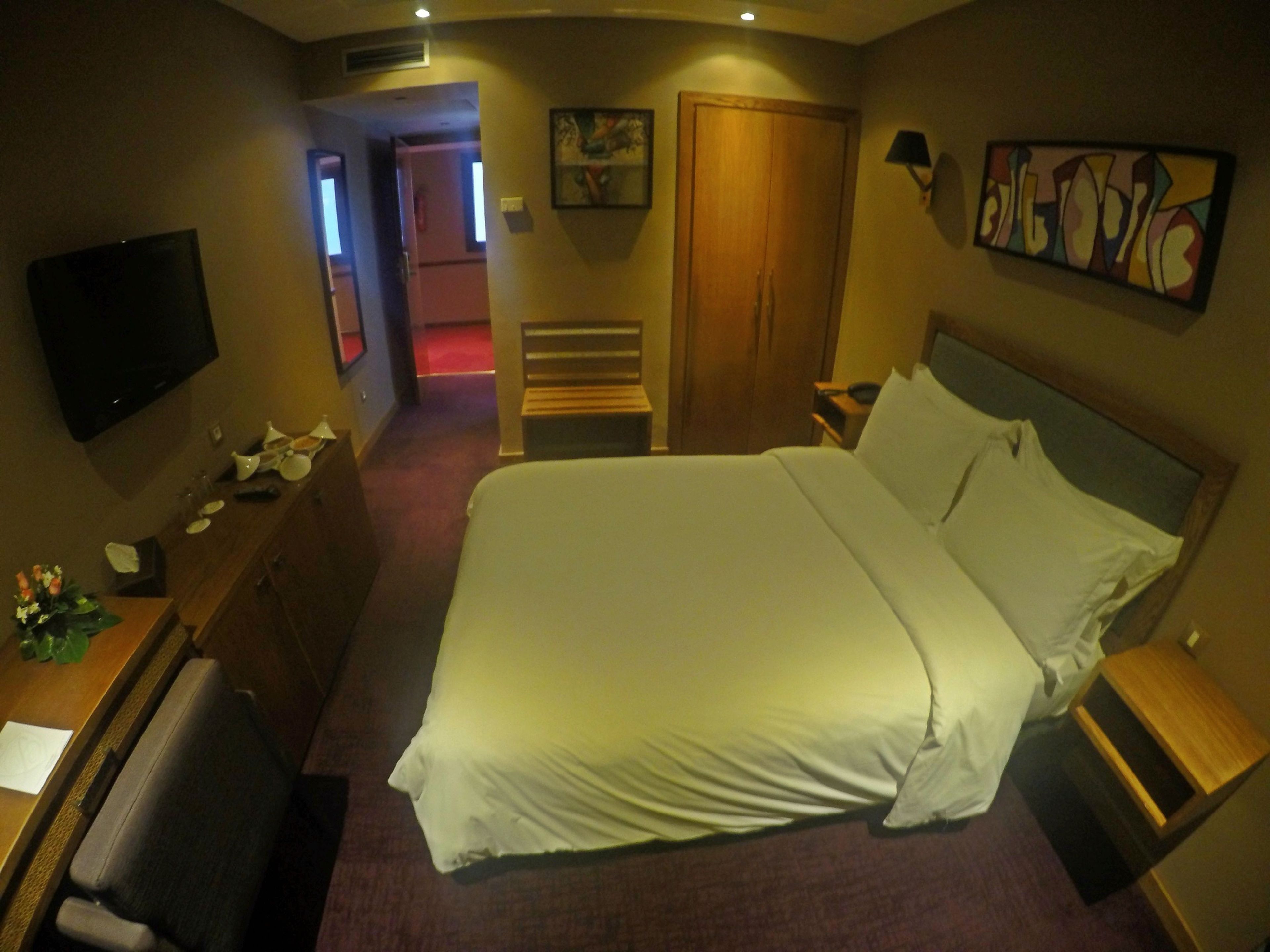 Quarto de hotel com uma cama, uma escrivaninha com cadeira, uma televisão e um quadro na parede.