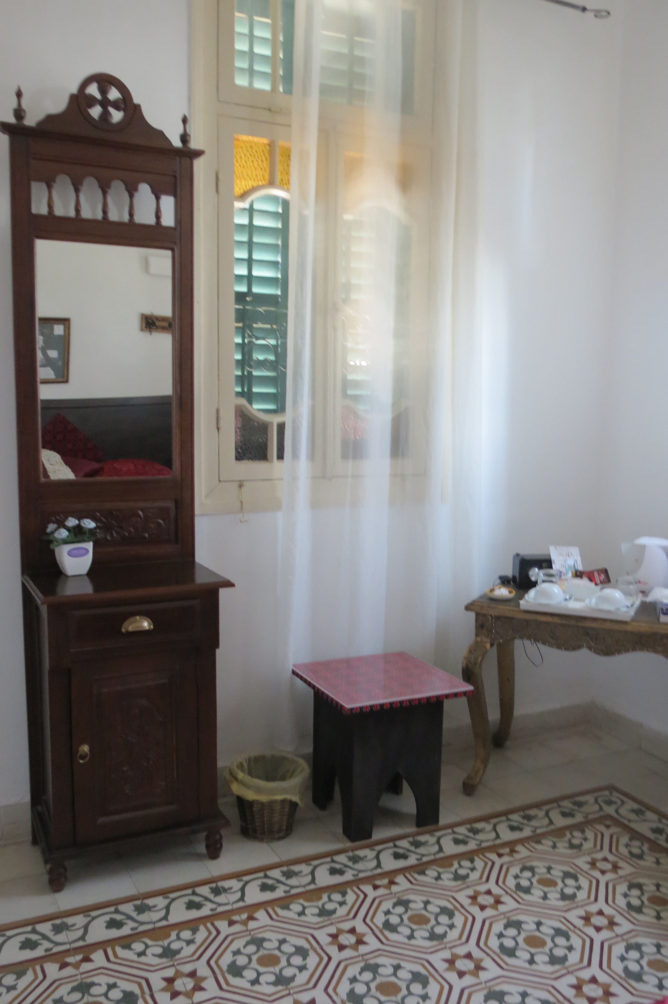 Quarto com cômoda de madeira, um banquinho pequeno, uma mesa com toalha branca e piso estampado.
