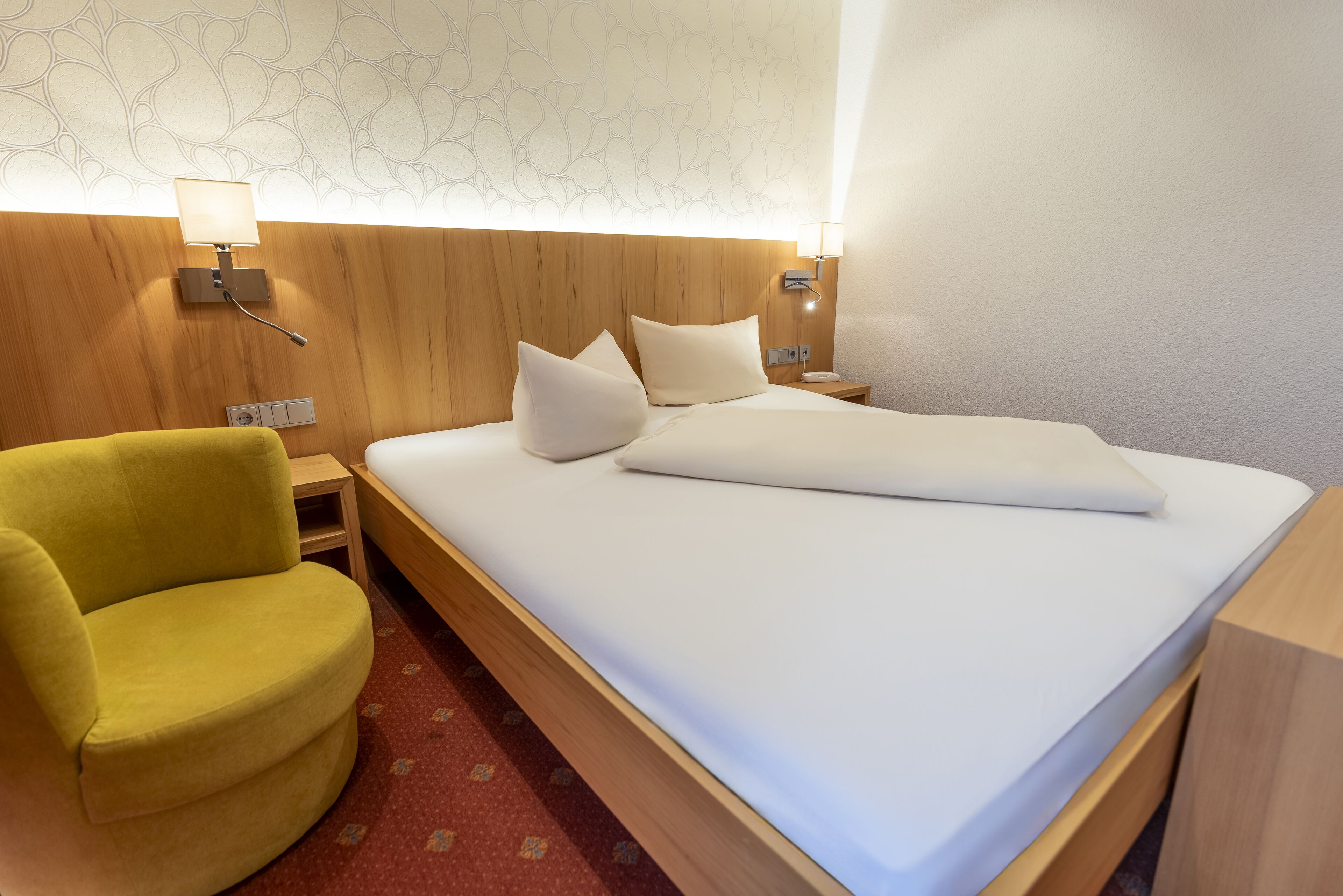 Quarto de hotel com cama, uma poltrona amarela e duas luminárias de cabeceira.