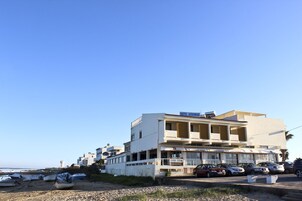Um hotel com vários andares, uma varanda e uma placa indicando ser hotel. Vários carros estão estacionados na frente, e barcos são visíveis na praia.