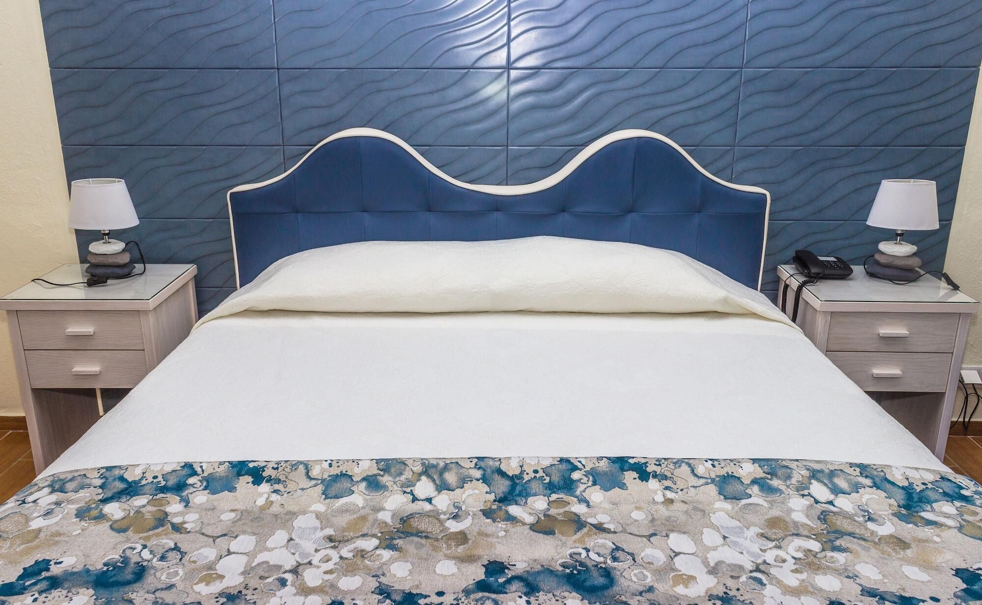 Uma cama com cabeceira azul, roupa de cama branca e manta estampada com flores azuis. Há duas mesas de cabeceira, cada uma com uma luminária e um telefone.