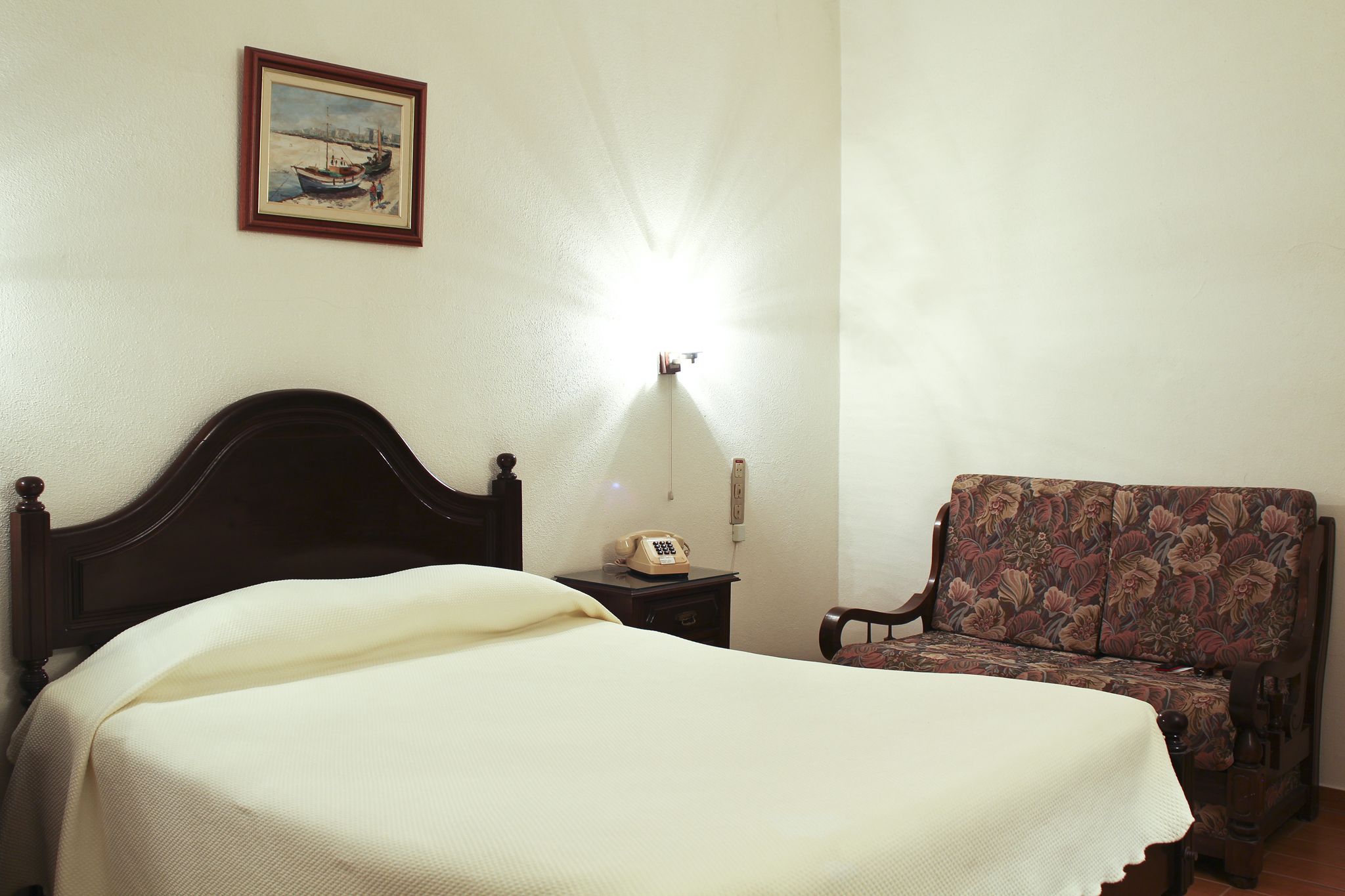 Quarto com cama, mesa de cabeceira com telefone, uma cadeira e luminária fixada na parede.