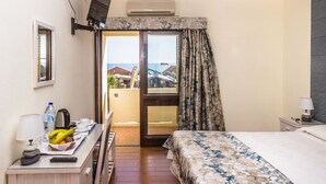 Quarto de hotel com cama, secretária com abajur, cadeira, mesa pequena com frutas e chaleira, e vista para o mar através de uma porta de vidro.